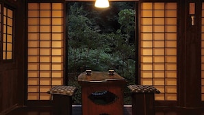 Um quarto japonês tradicional com tatames, uma mesa de madeira baixa com almofadas e vista para um jardim exuberante através de biombos shoji.