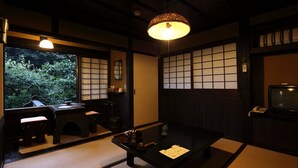 Um quarto japonês tradicional com tatames, uma mesa baixa com bandeja, luminária pendente e uma televisão.