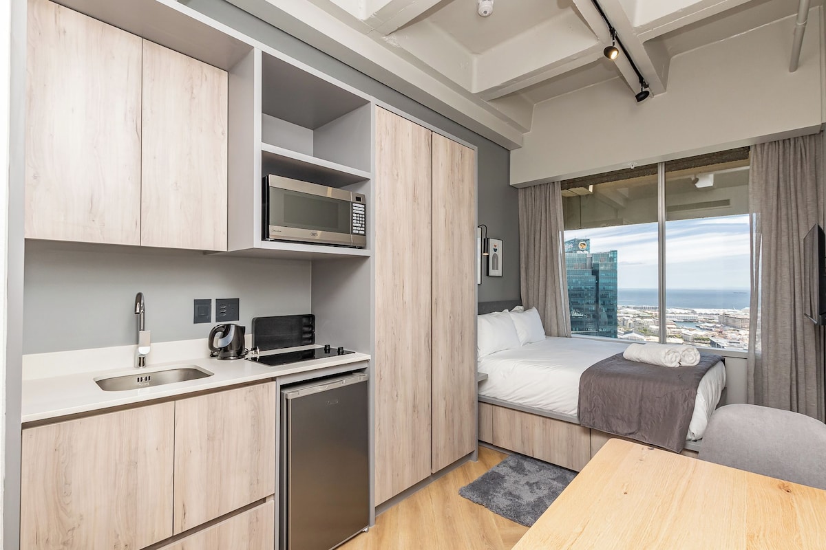Quarto de hotel moderno com cozinha compacta, cama com vista e uma janela grande.