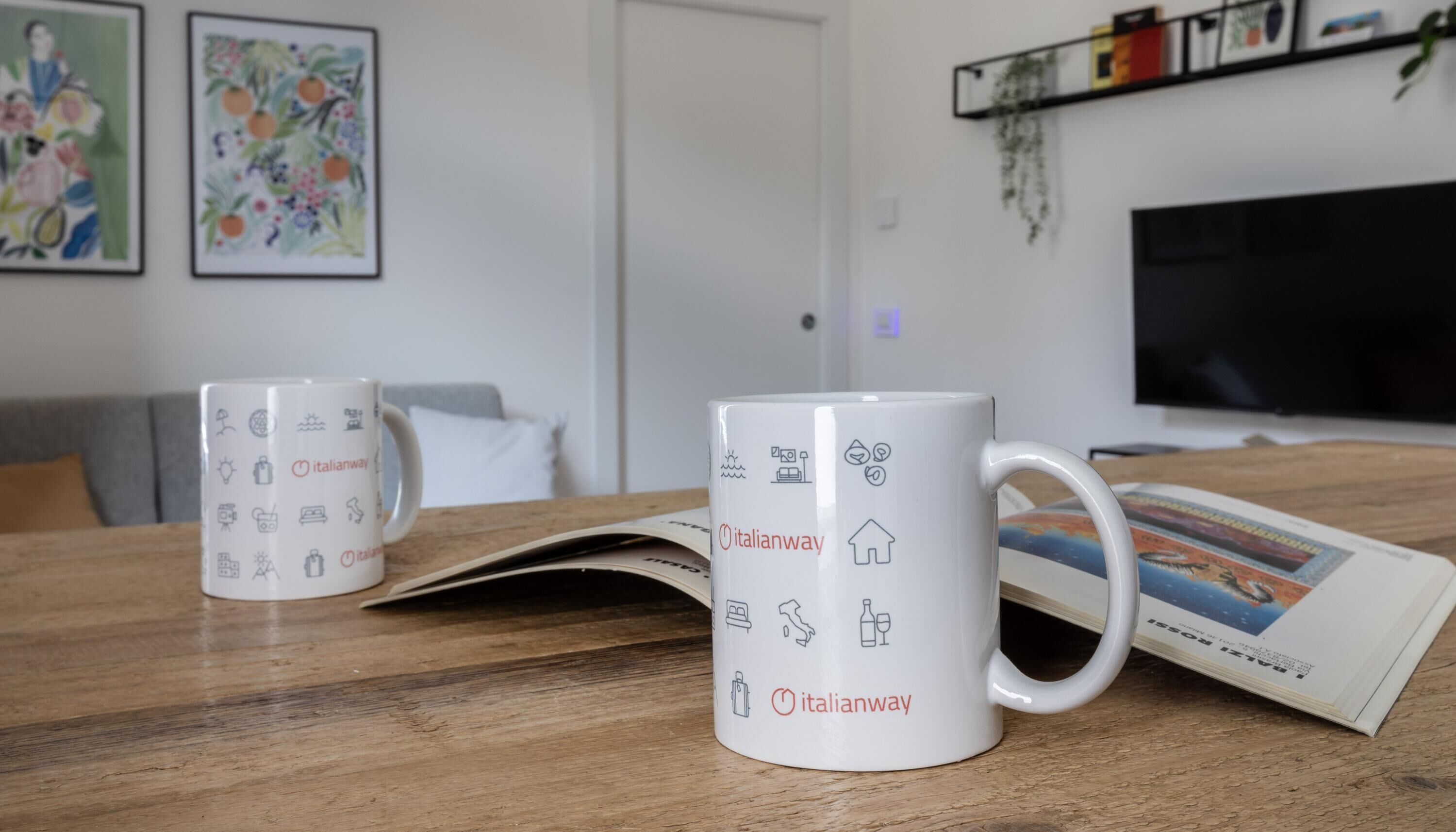 Duas canecas brancas com a marca Italianway sobre uma mesa de madeira, uma revista e uma televisão em uma sala de estar.