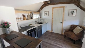 Uma cozinha compacta com forno embutido, pia e torradeira. Há uma mesa de madeira com um vaso de flores e uma poltrona marrom com almofada.