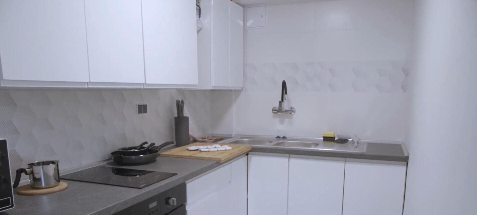 Cozinha com armários brancos, pia de aço inoxidável, fogão por indução preto e tábua de corte de madeira.