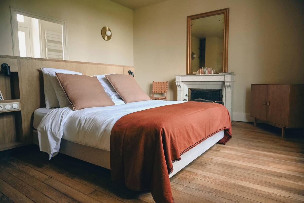 Uma cama bem arrumada com edredom branco e castanho, chão de madeira, uma cadeira de madeira pequena, lareira e um espelho na parede.
