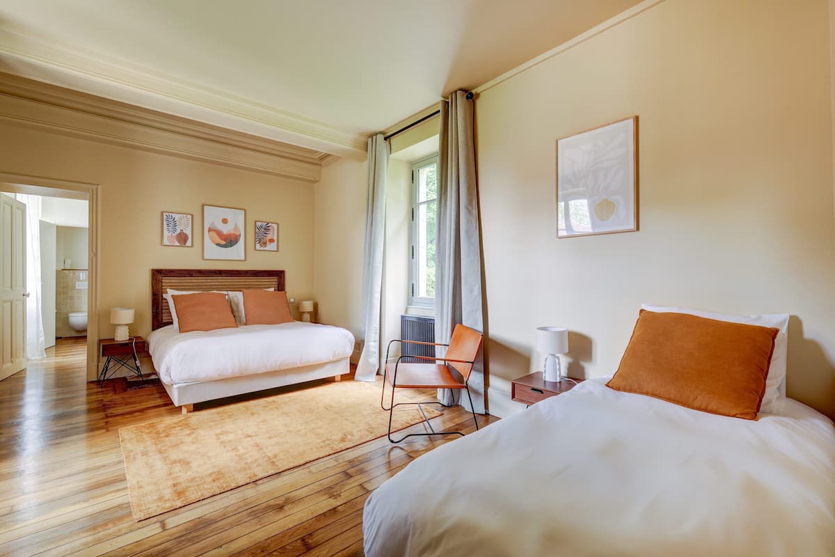 Quarto de hotel com duas camas, piso de madeira, uma cadeira, radiador e obras de arte emolduradas nas paredes.