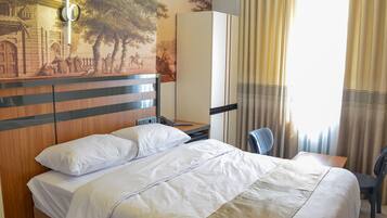 Cama bem arrumada com lençóis brancos e manta bege, cabeceira de madeira, luminária na parede e uma janela grande com cortinas.