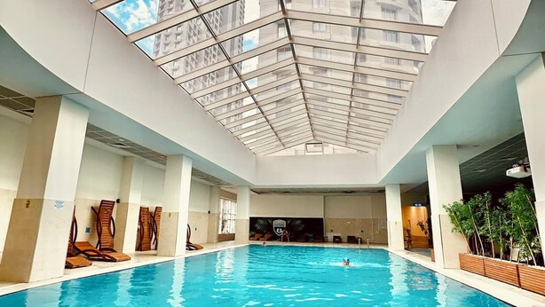 Piscina interna com teto de vidro, cadeiras de descanso e vista para edifícios altos.