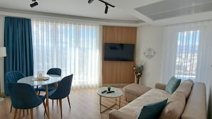 Uma sala de estar moderna com uma mesa de jantar redonda, cadeiras azuis, um sofá bege e uma TV fixada na parede.
