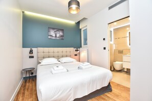 Quarto de hotel moderno com uma cama grande, uma pequena mesa de cabeceira, uma luminária fixada na parede e uma moldura com uma imagem sobre a cama.
