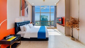 Quarto de hotel moderno com uma cama grande, televisão, planta em vaso e vista da cidade através de amplas janelas.