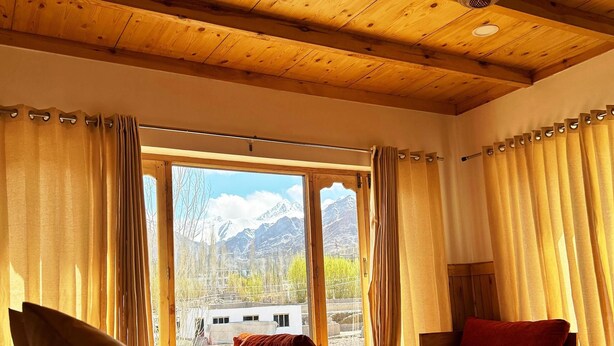 Teto de madeira, cama com almofadas, janela com cortinas e vista para montanhas.