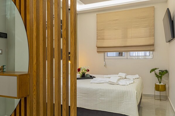Quarto de hotel com uma cama, mesa de cabeceira com um vaso de flores, janela com persiana bege e TV fixada na parede.