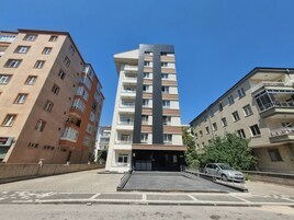 Edifício residencial com vários andares, entrada central, varandas e área de estacionamento.
