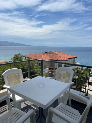 Uma varanda com mesa e cadeiras brancas, com vista para a costa e um edifício com telhado vermelho ao fundo.