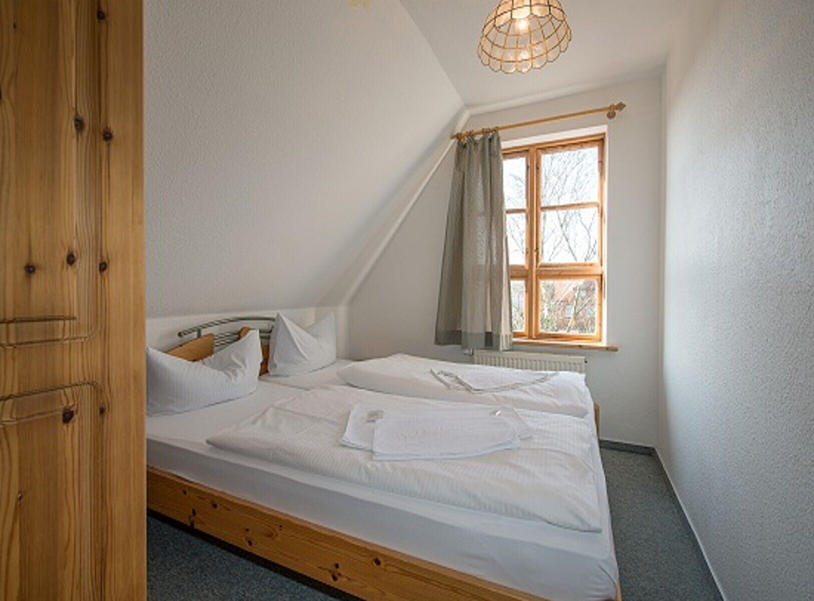 Quarto com estrutura de cama de madeira, roupa de cama branca, janela com cortinas e porta de madeira.