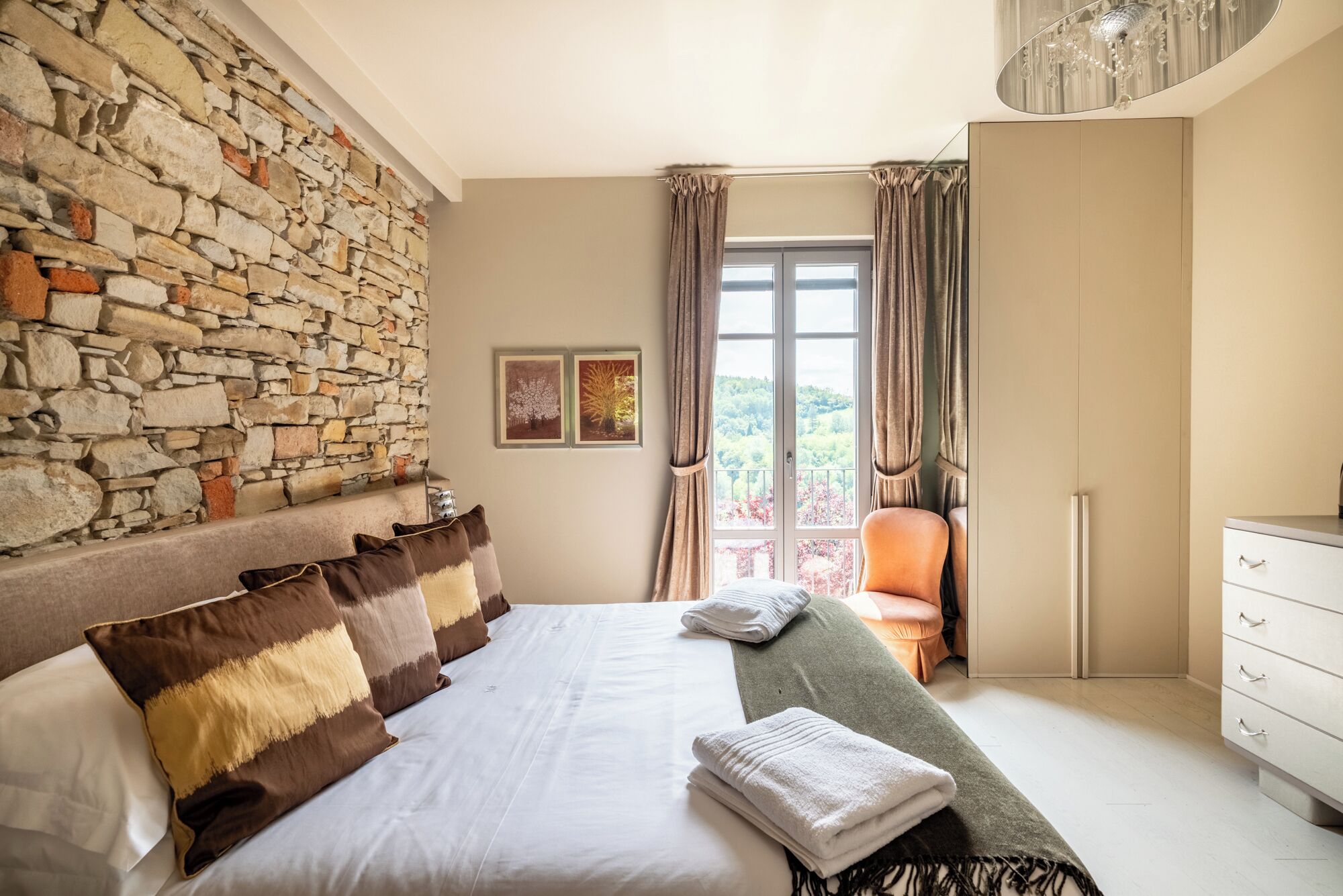 Quarto com parede de pedra, cama com várias almofadas, janela com cortinas, uma cadeira e um aparador.