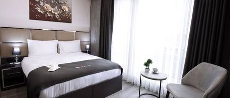 Quarto de hotel moderno com uma cama grande, mesas de cabeceira, uma pequena mesa redonda, uma cadeira e um objeto decorativo na parede.