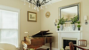 Uma sala de estar com um piano de cauda, lareira, lustre e um espelho grande.