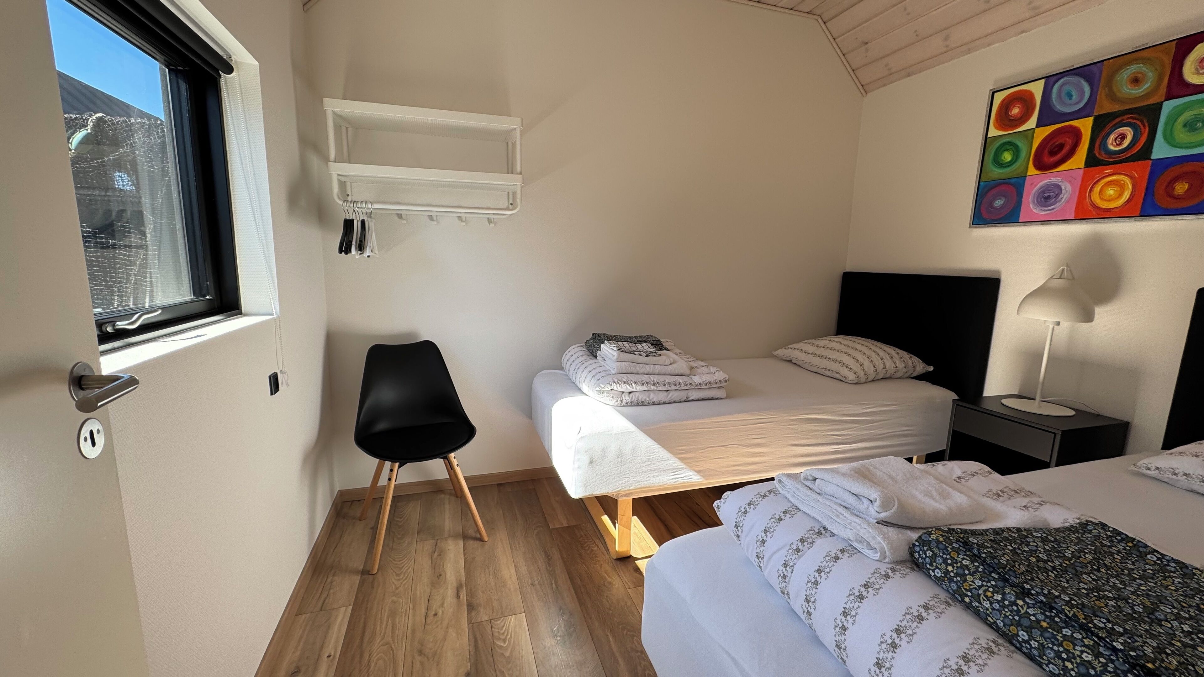 Quarto de hotel compacto com uma cama de solteiro, uma pequena mesa de cabeceira com abajur, uma cadeira e uma prateleira na parede.