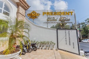 Exterior do Hotel President, com destaque para a placa com o nome do hotel, entrada curva, plantas em vasos e uma motocicleta estacionada.