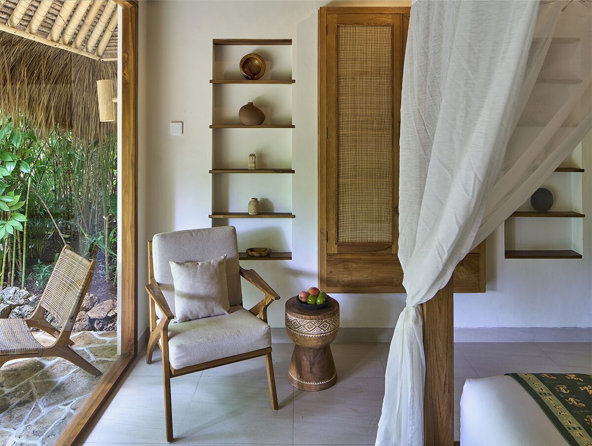 Quarto com uma cadeira de madeira, uma mesa redonda pequena com frutas, uma estante com objetos decorativos e uma janela com vista para o verde.
