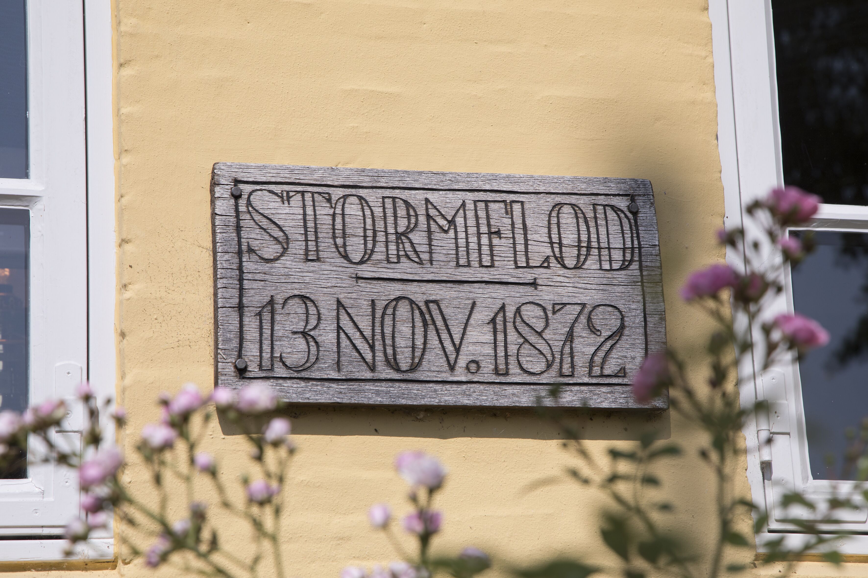 Uma placa de madeira com o texto 'STORMFLOOD 13 NOV. 1872' fixada em uma parede amarela.