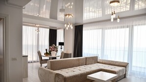 Uma sala de estar espaçosa com um amplo sofá seccional bege, uma mesa de centro branca e um lustre. Ao fundo, há uma área de jantar com uma mesa posta para dois e um vaso de flores.