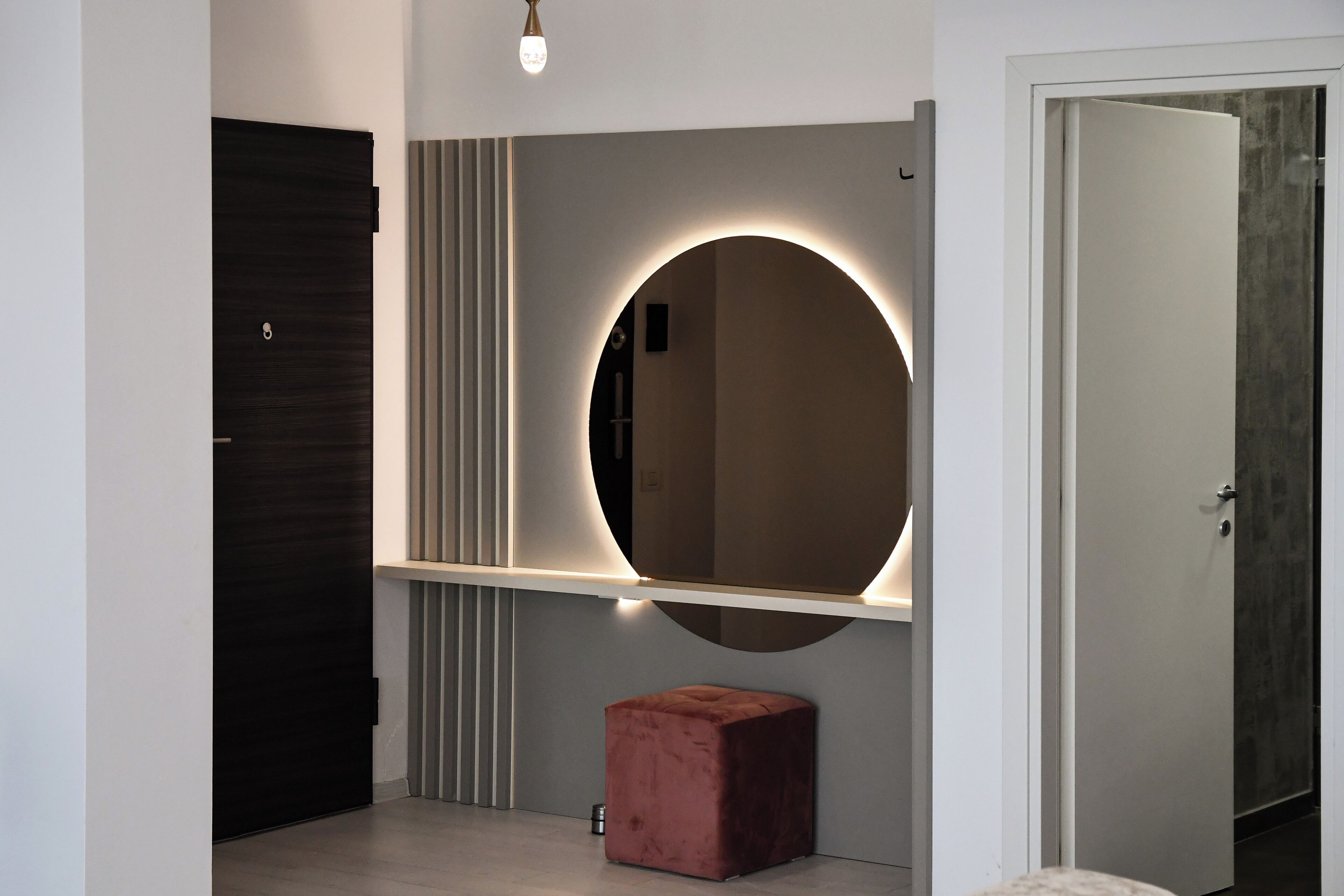Um interior moderno com uma porta escura, um espelho redondo com um brilho luminoso e um banquinho vermelho.