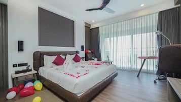 Quarto de hotel moderno com uma cama grande, uma escrivaninha, uma cadeira, um ventilador de teto e vista para o exterior através de amplas janelas.