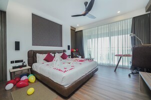 Quarto de hotel moderno com uma cama grande, uma escrivaninha, uma cadeira, um ventilador de teto e vista para o exterior através de amplas janelas.