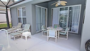 Uma área de pátio externa com mobiliário branco, incluindo cadeiras e uma mesa, e um ventilador de teto.