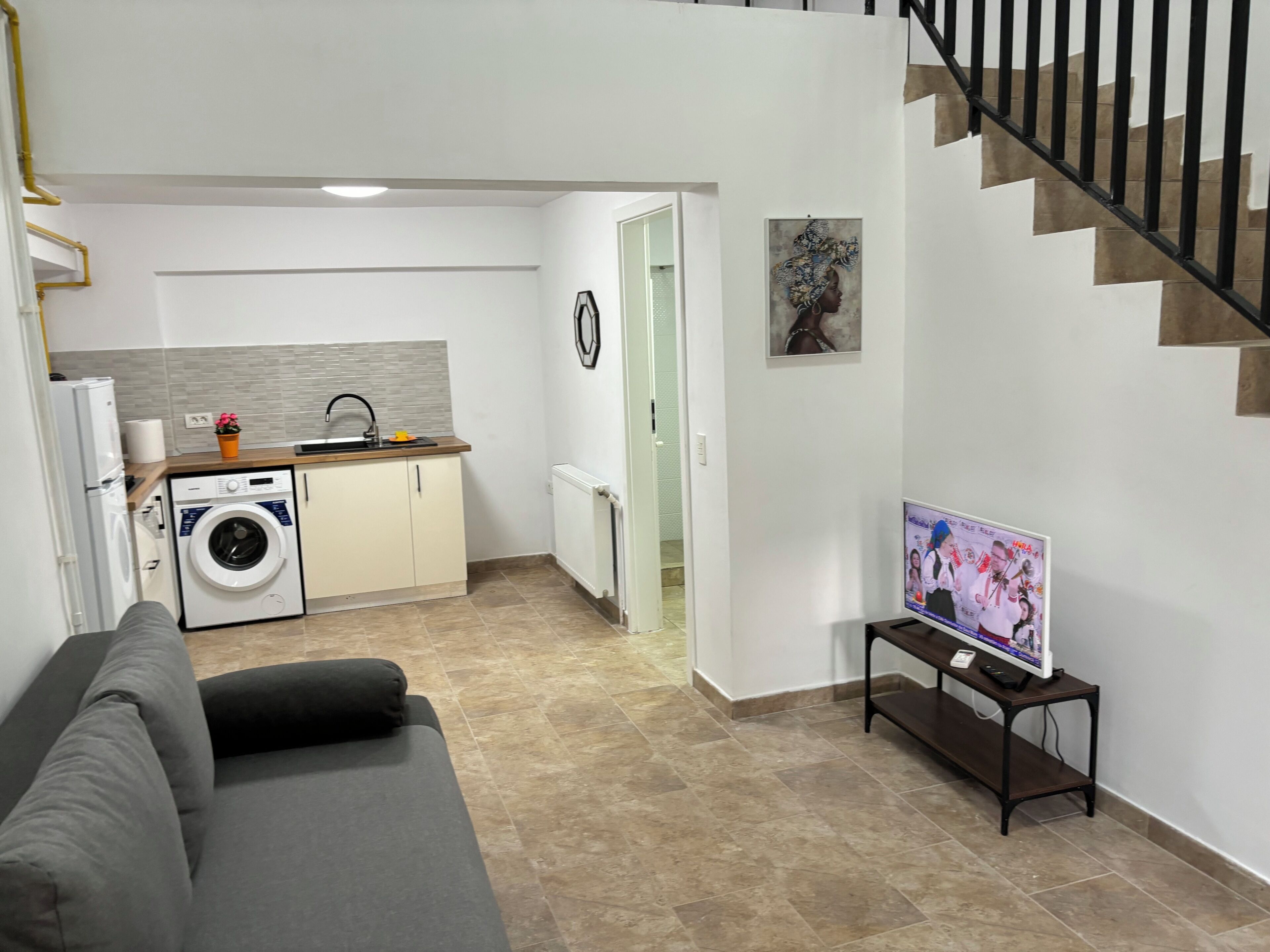Uma sala de estar moderna com um sofá cinzento, cozinha com máquina de lavar roupa, mesa de jantar e escadas.