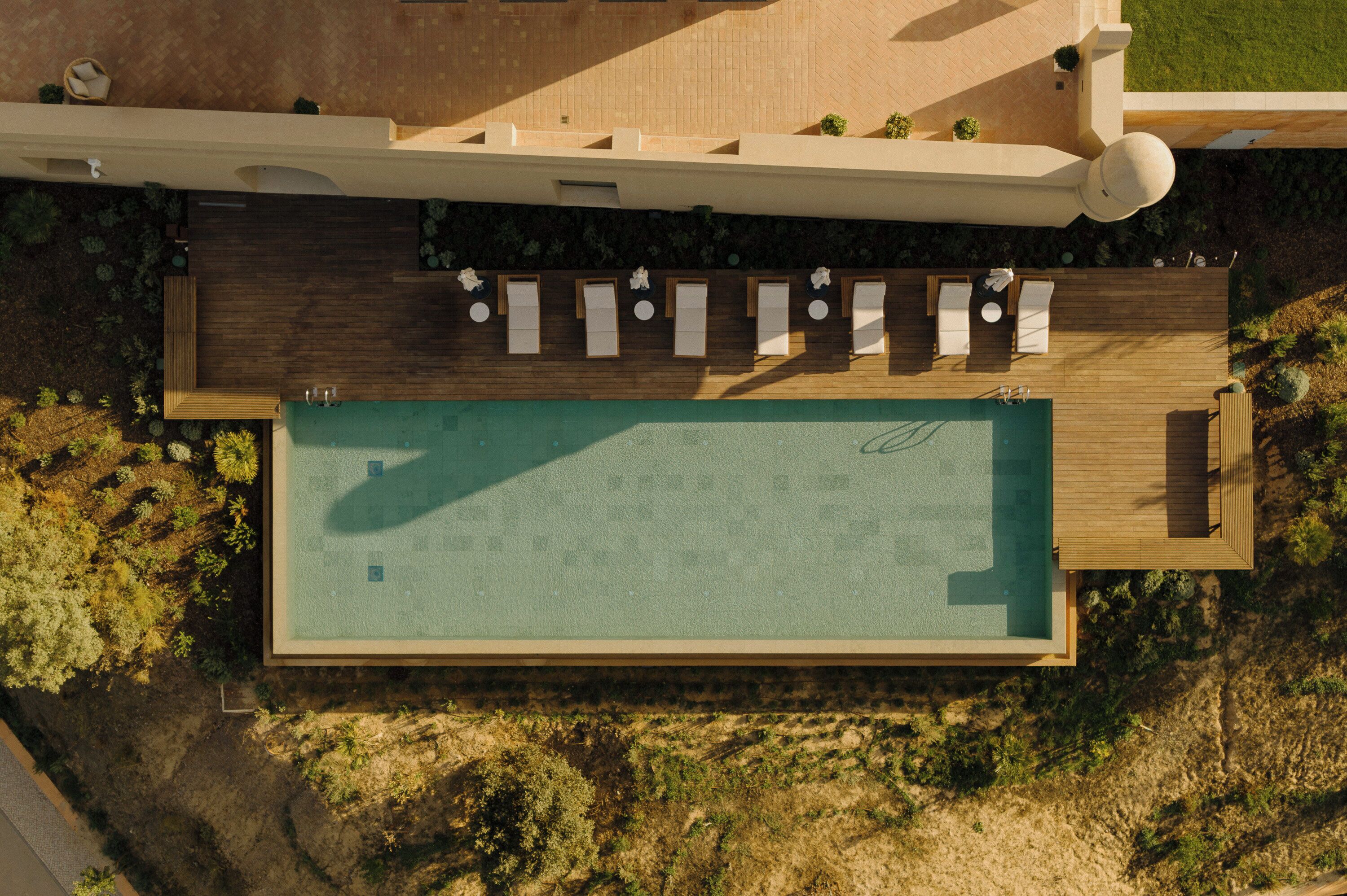 Vista aérea de uma piscina retangular com um deck de madeira ao redor, equipada com várias espreguiçadeiras e uma pequena mesa.