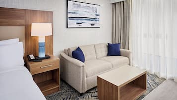 Quarto de hotel com uma cama, mesa de cabeceira com luminária, sofá com almofadas azuis, mesa de madeira pequena e um quadro na parede.