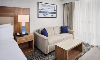 Quarto de hotel com uma cama, mesa de cabeceira com luminária, sofá com almofadas azuis, mesa de madeira pequena e um quadro na parede.