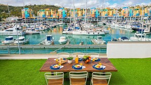 Mesa de jantar com café da manhã em terraço com vista para uma marina, com edifícios coloridos e barcos.