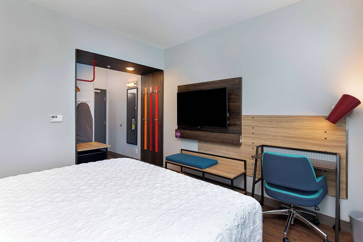 Quarto de hotel com uma cama, uma escrivaninha com cadeira, uma TV fixada na parede e um abajur vermelho.