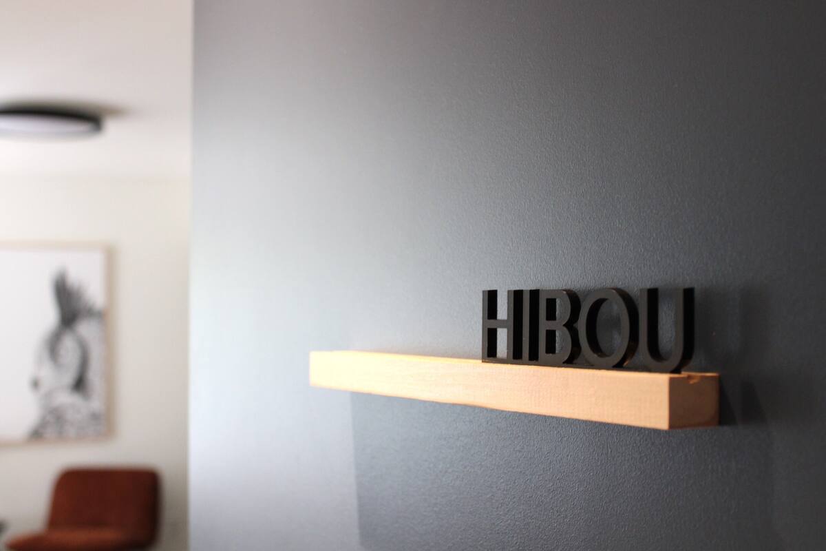 Quarto minimalista com uma prateleira de madeira exibindo a palavra "HIBOU" em uma parede cinza.