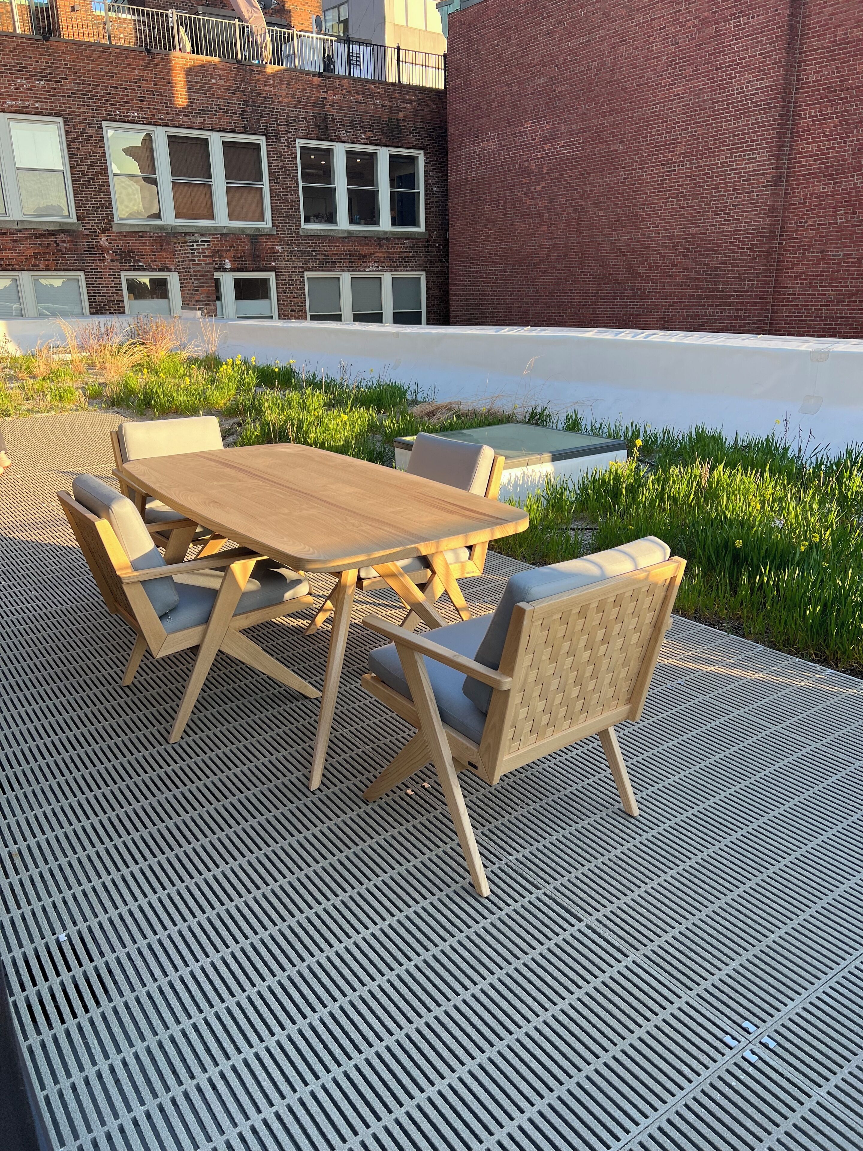 Um pátio externo com mesa e cadeiras de madeira, cercado por piso de grade metálica e vegetação.