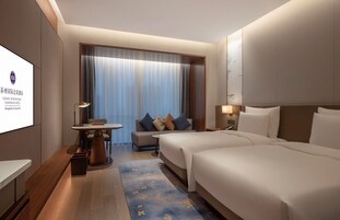 Quarto de hotel com cama, sofá, escrivaninha e televisão exibindo informações do hotel.