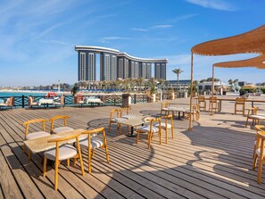 Um deck de madeira com mobiliário para áreas externas, com vista para uma praia e um hotel alto.