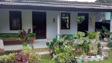 Uma casa com telhado de cerâmica, varanda frontal com um banco e diversos vasos de plantas na varanda e no jardim.