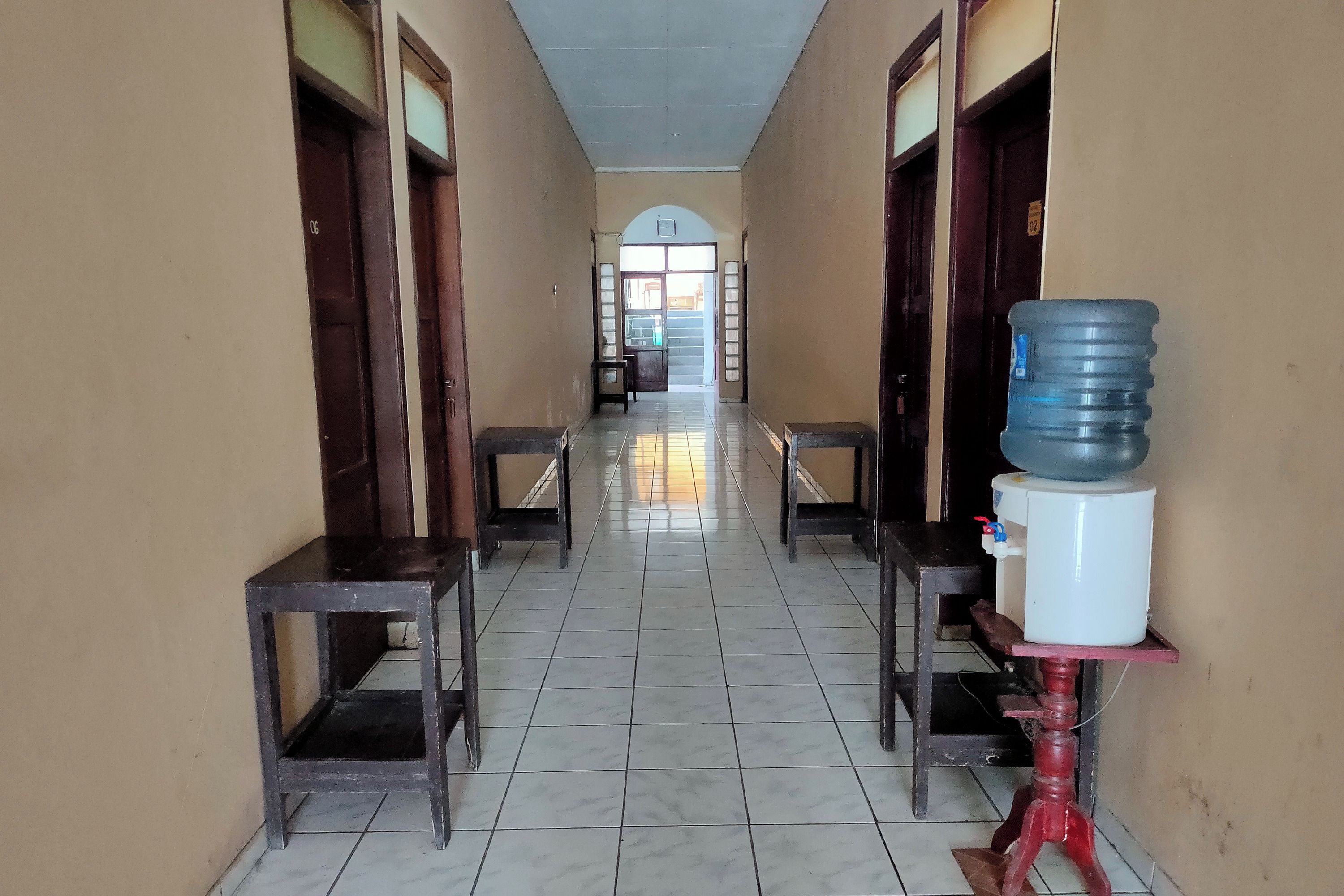 Um corredor com piso de azulejos, dois bancos de madeira e um bebedouro à direita.