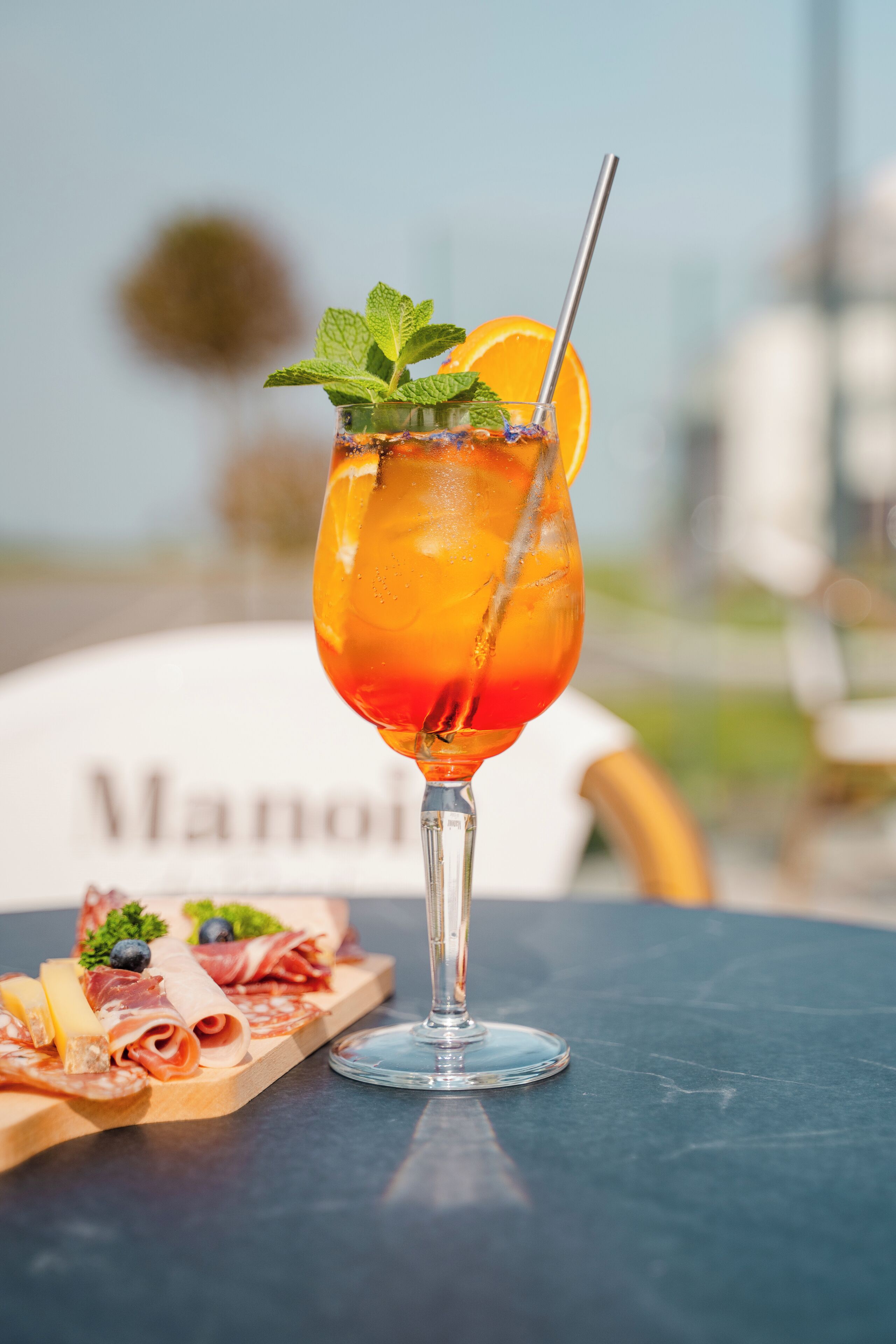 Um copo de cocktail com um ramo de hortelã e uma fatia de laranja, uma tábua de madeira com diversos carnes e queijos, e uma mesa com uma cadeira ao fundo.