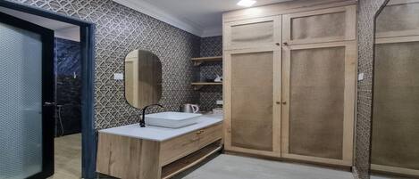 Casa de banho com toucador de madeira, lavatório branco e espelho. Tem zona de duche com porta de vidro e chão em mosaicos.