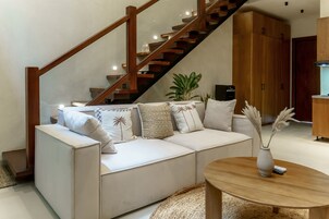 Uma sala de estar moderna com escada de madeira, um sofá branco com almofadas estampadas, uma mesa de centro de madeira e uma planta em vaso.