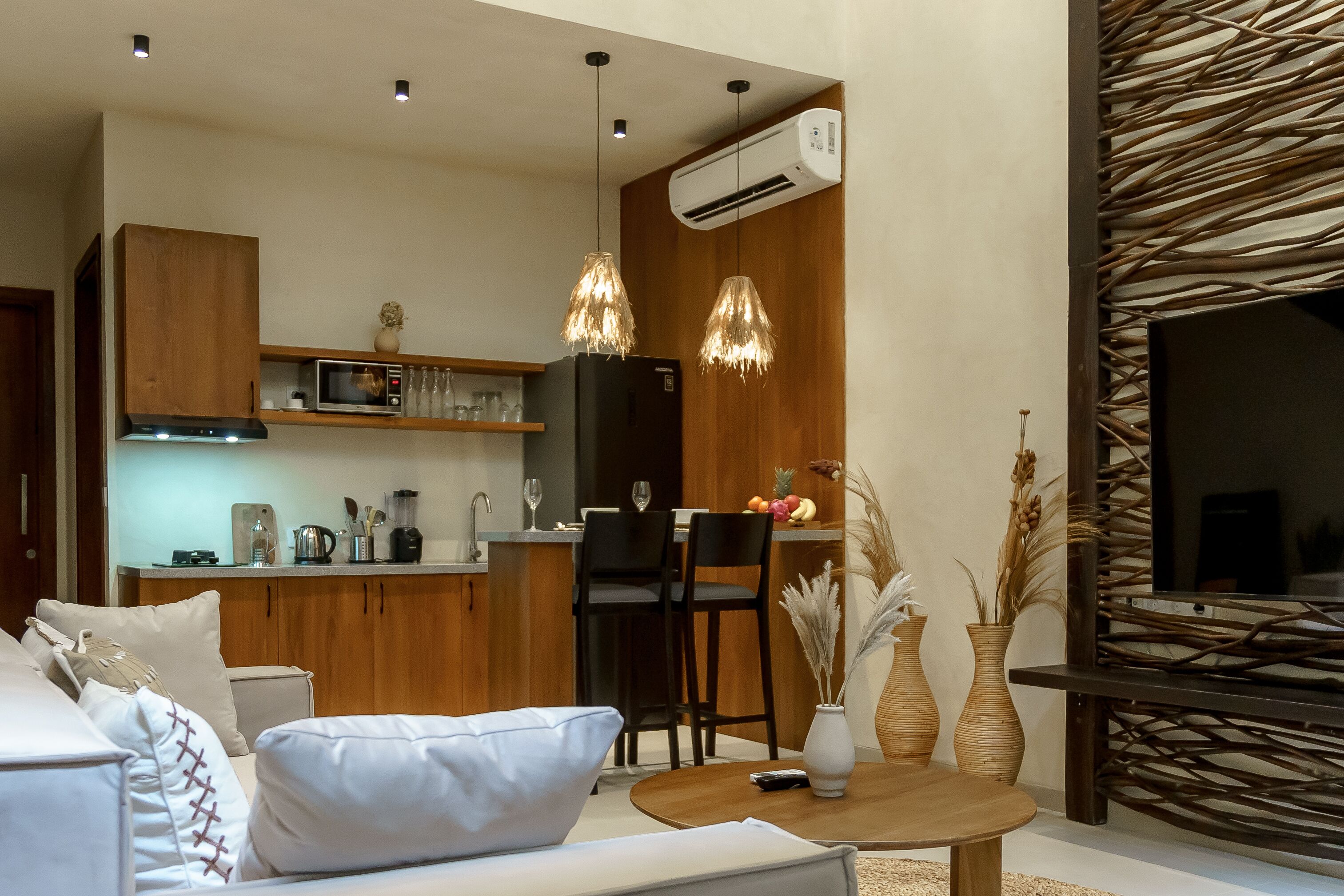 Uma sala de estar moderna com um sofá, mesa de centro e vasos decorativos. Uma cozinha com geladeira, micro-ondas e mesa de jantar é visível ao fundo.