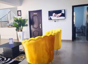 Uma área de recepção com uma porta de madeira escura, uma televisão fixada na parede e dois assentos amarelos.