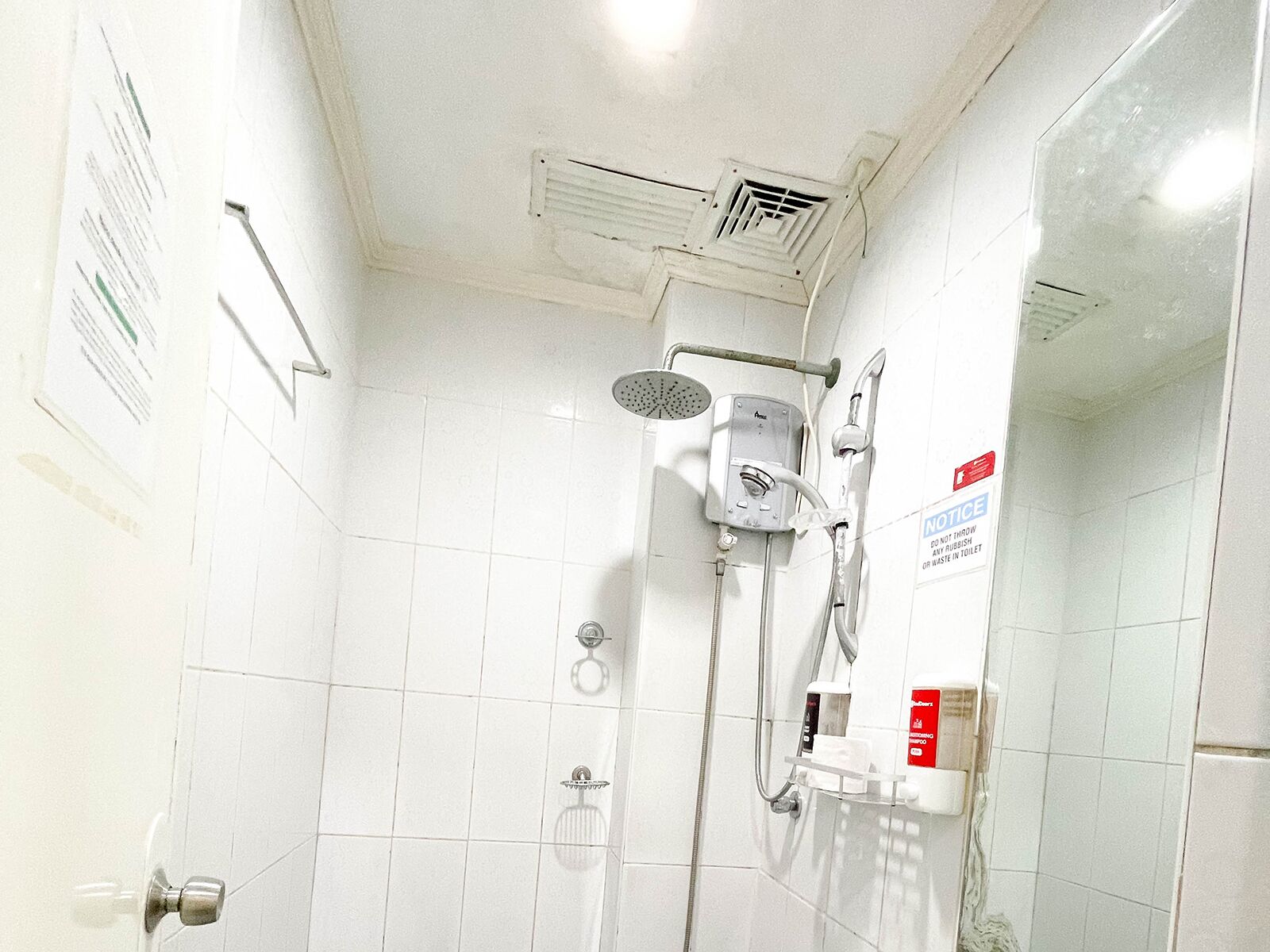Um chuveiro pequeno com azulejos brancos, um chuveiro manual, um painel de controle na parede e uma placa de aviso vermelha.