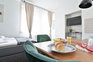 Quarto de hotel moderno com cama, mesa de café da manhã com croissant e suco, televisão e janela com cortinas.