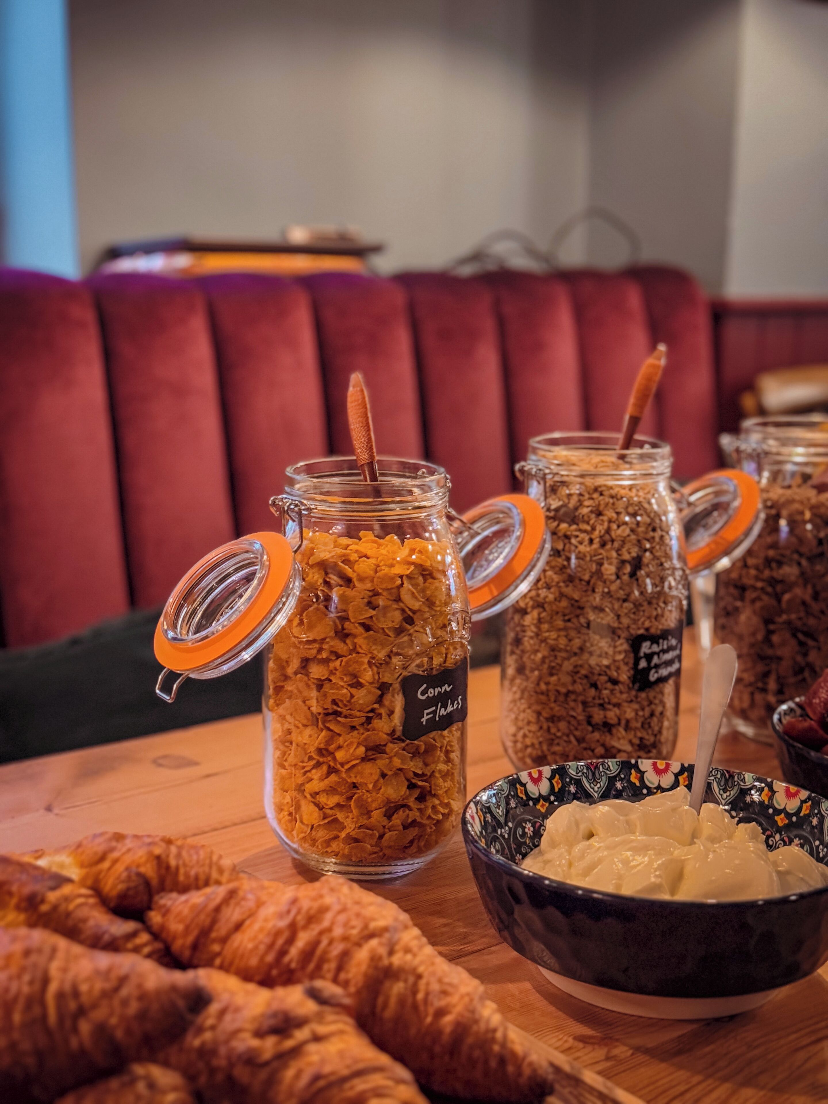 Um buffet de café da manhã com potes de cereais, um prato de iogurte e croissants em uma mesa de madeira.